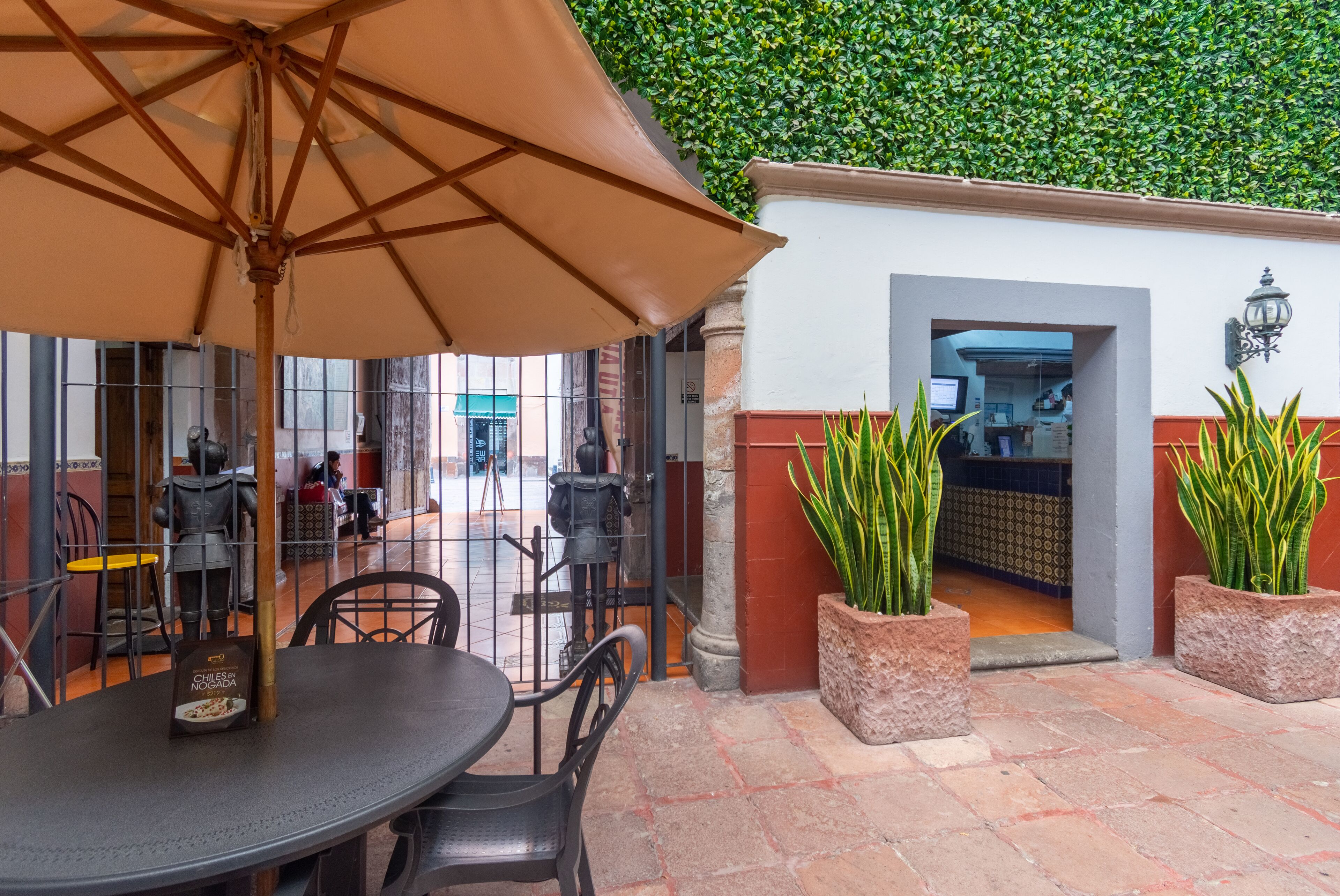 Área de pátio externa com mesa e cadeiras sob um guarda-sol grande, plantas em vasos e uma parede com design de folhagem verde.