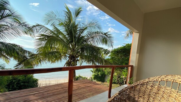 Varanda com cadeira de vime e mesa pequena, com vista para uma praia e palmeiras.