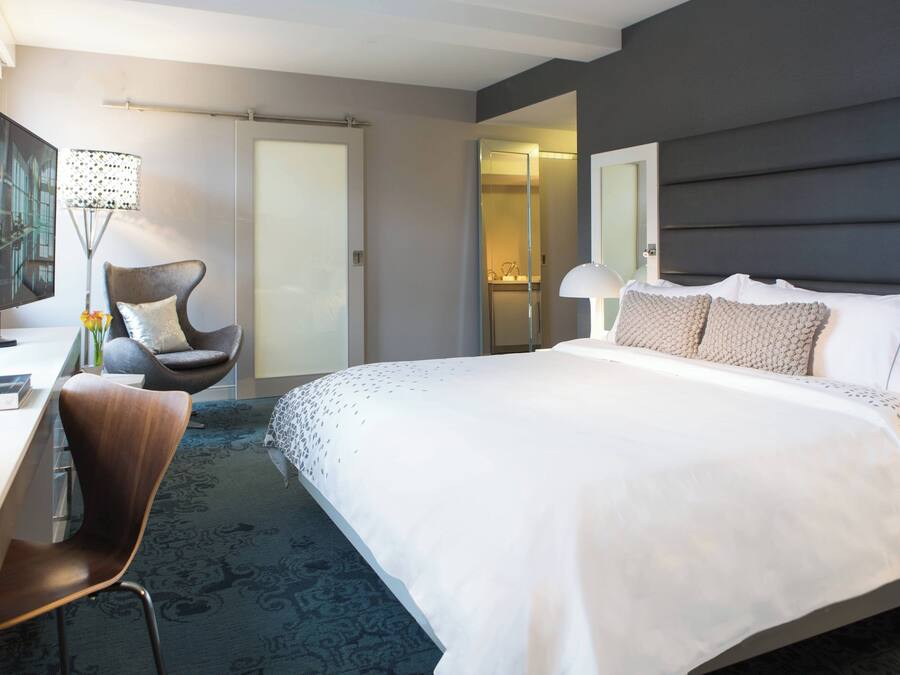 Quarto de hotel moderno com uma cama grande, uma escrivaninha com cadeira, uma luminária de mesa e um espelho grande.