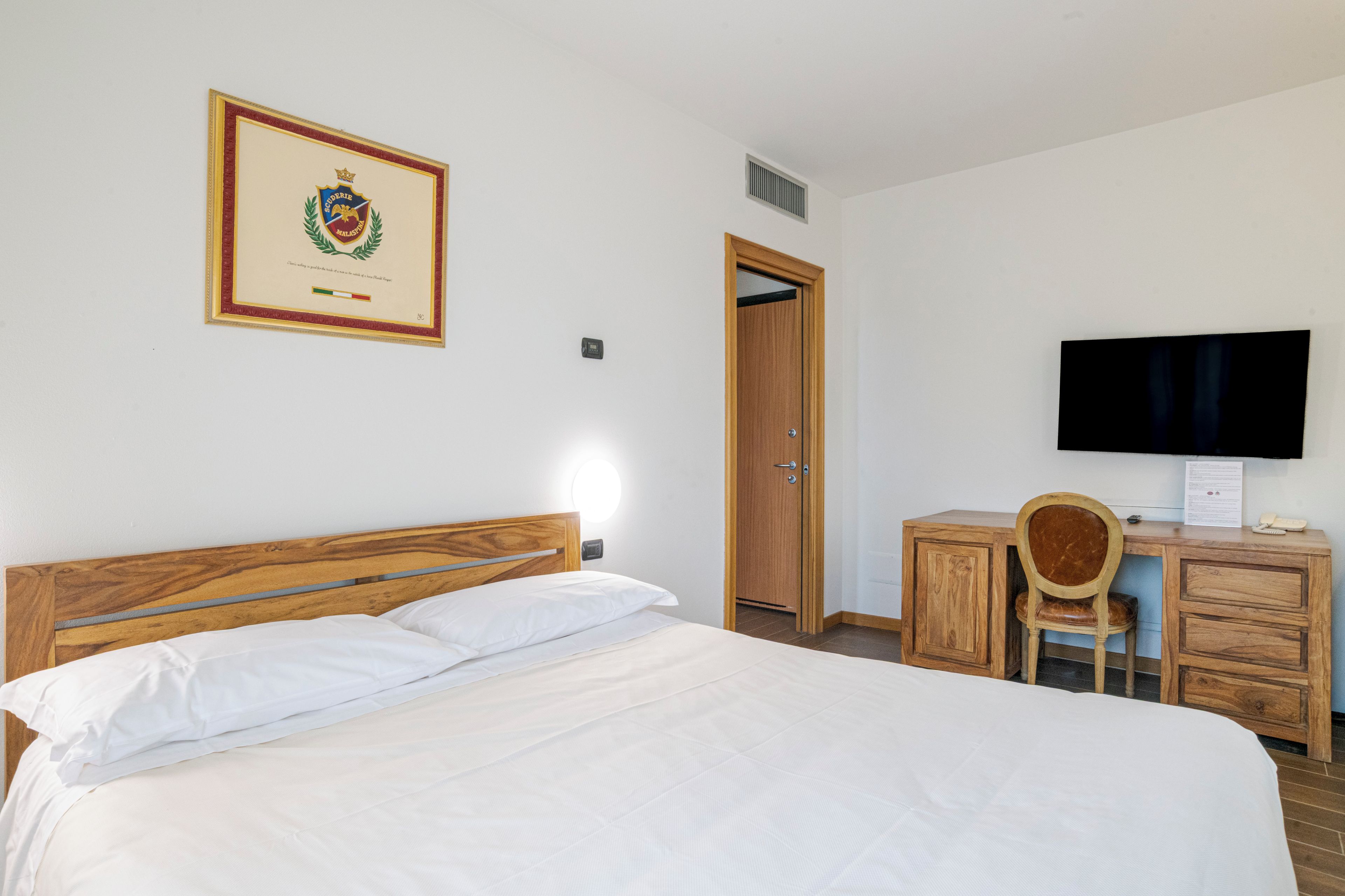 Quarto de hotel com cama, escrivaninha, cadeira, televisão e porta.