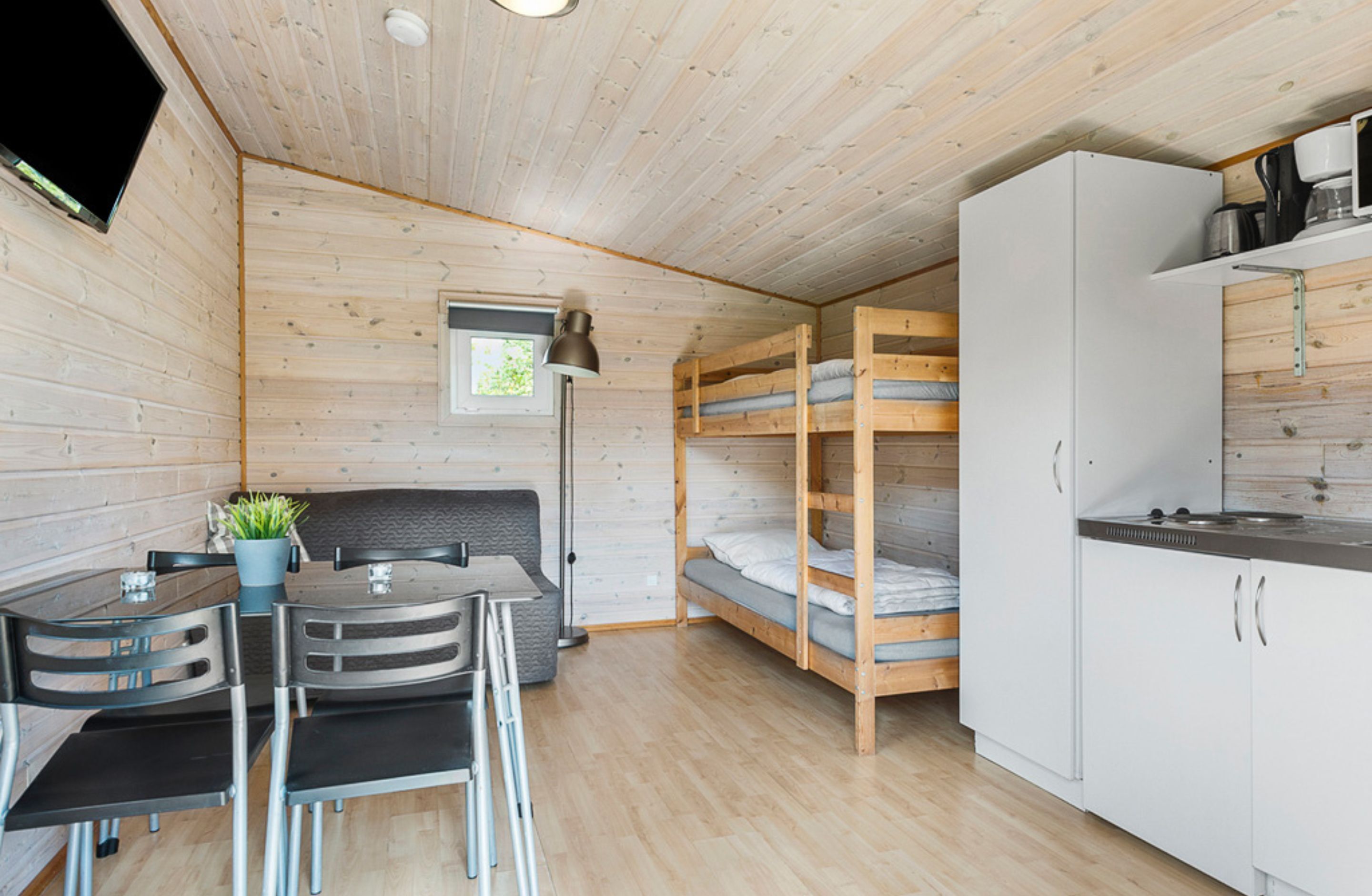 Quarto compacto com beliches, pequeno espaço de cozinha, área de jantar com mesa e cadeiras, e uma janela.