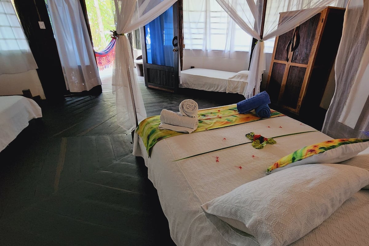 Quarto com duas camas, cada uma com roupa de cama branca e dossel. Há um tapete colorido no chão e uma toalha enrolada em uma das camas.