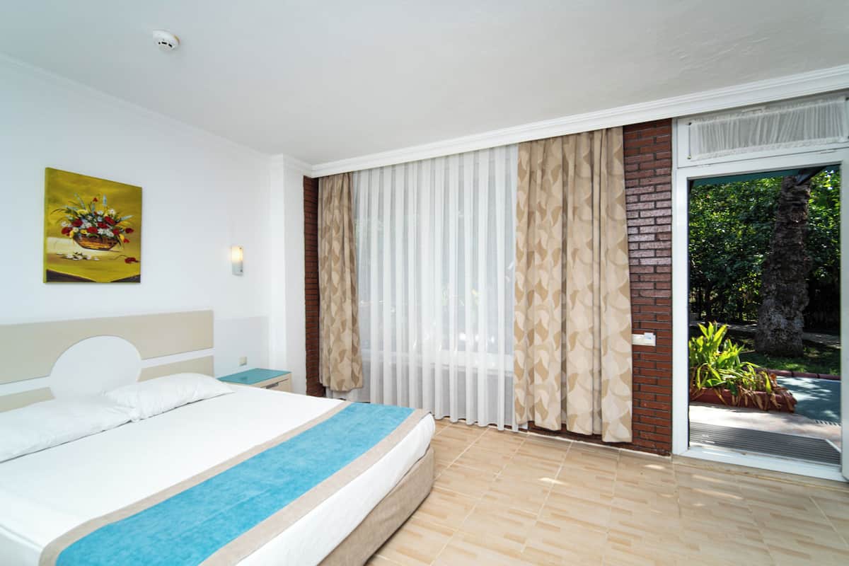 Um quarto com cama, mesa de cabeceira, quadro, janela com cortinas e vista para o verde lá fora.