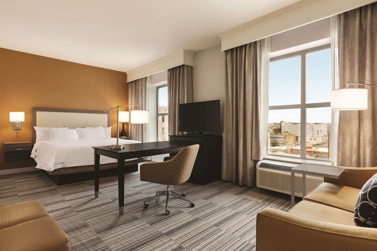 Quarto de hotel com cama, escrivaninha, cadeira, televisão e amplas janelas com cortinas.