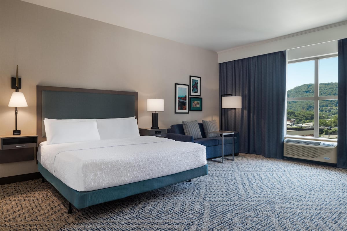 Quarto de hotel com uma cama grande, duas mesas de cabeceira com luminárias, uma pequena escrivaninha, um sofá e uma janela ampla com vista para o exterior.