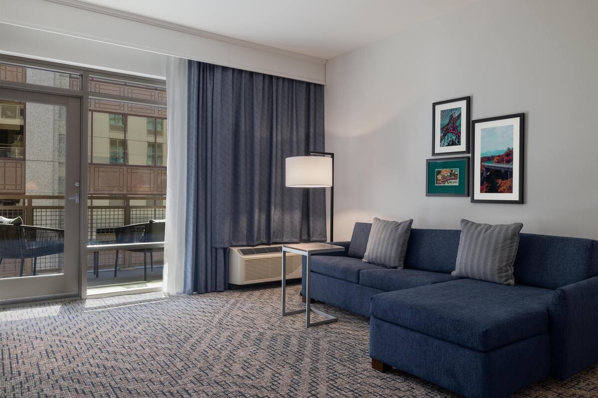 Quarto de hotel moderno com um sofá azul, uma mesinha, um abajur e uma janela grande com vista para um edifício.