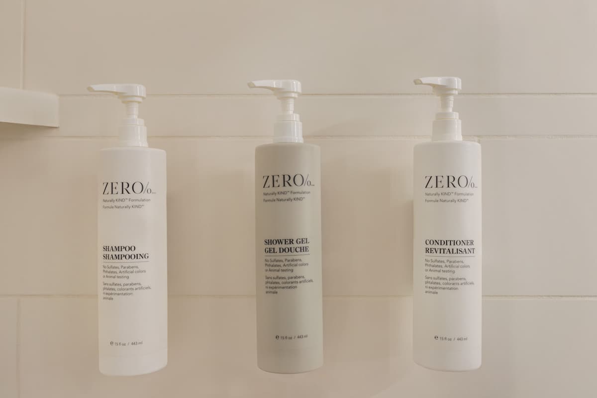 Três embalagens de shampoo, gel de banho e condicionador ZERO%, sobre uma parede de azulejos brancos.