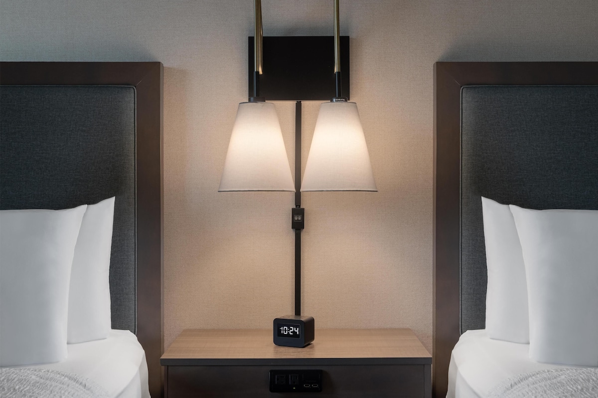 Quarto de hotel com duas camas, uma mesa de cabeceira com relógio digital e uma luminária fixada na parede.