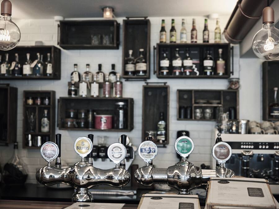 Uma área de bar com várias torneiras identificadas por nomes de cerveja e uma variedade de garrafas nas prateleiras ao fundo.