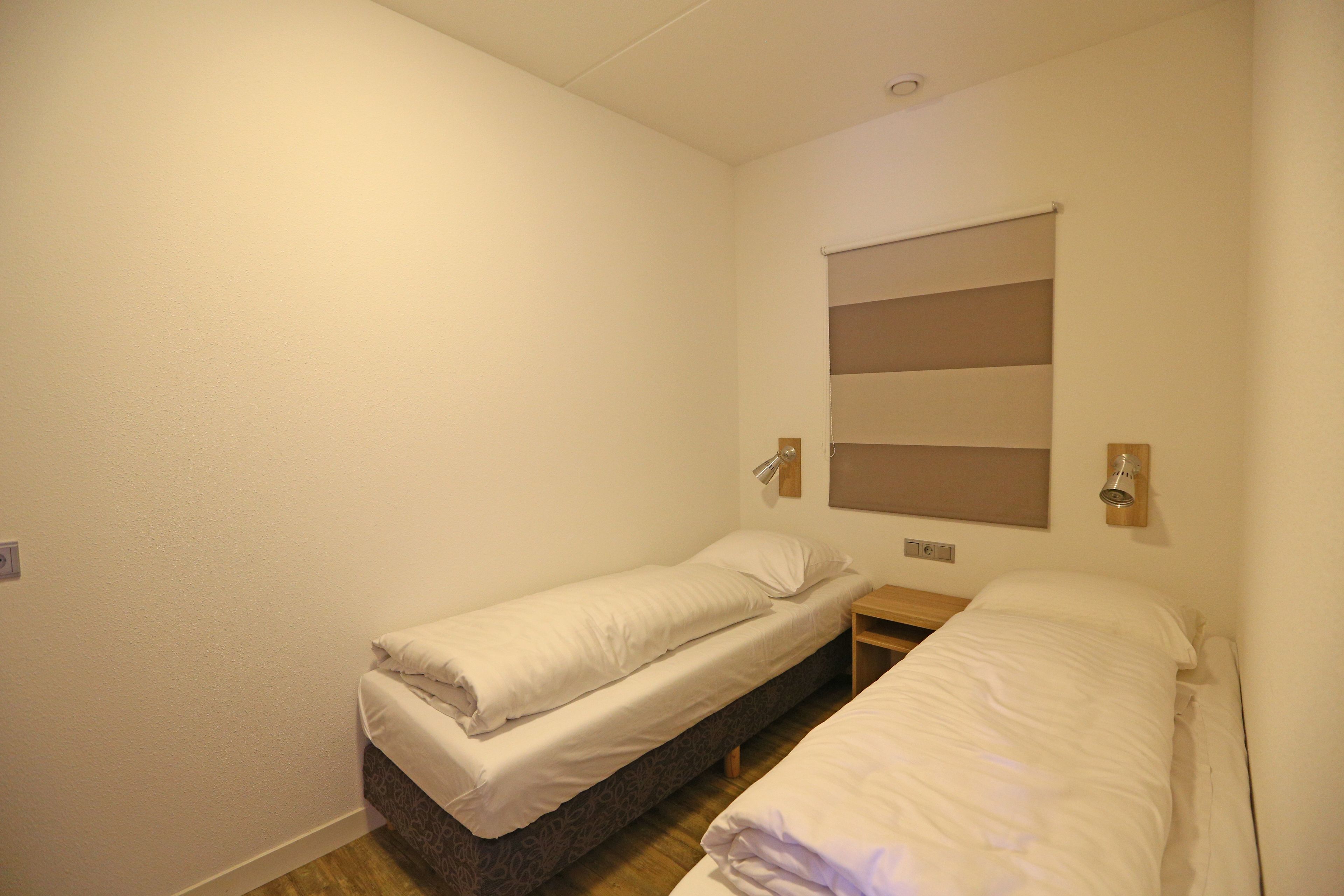 Acomodação modesta, com um quarto único e duas camas individuais, mesa de cabeceira de madeira, luminária na parede e janela com persianas.