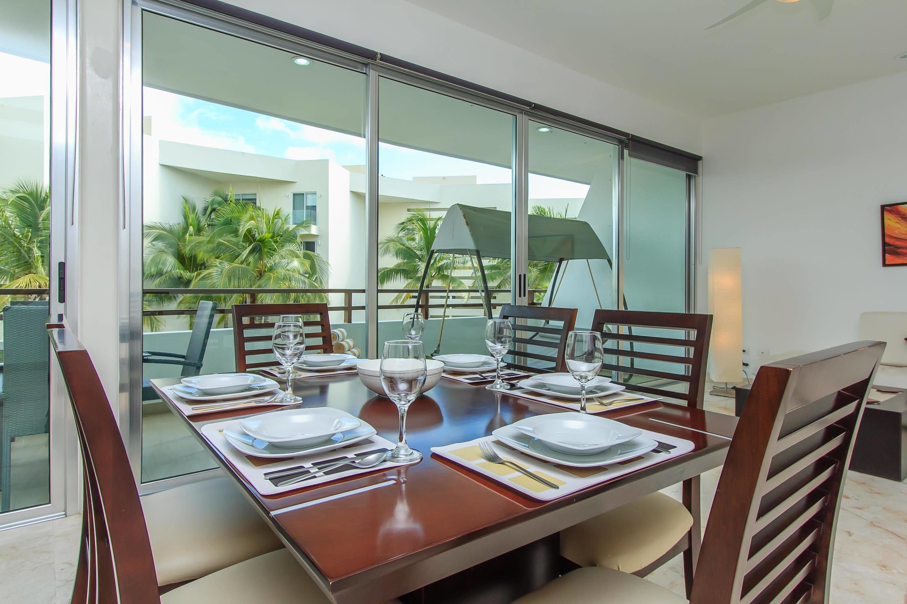 Uma sala de jantar com uma mesa posta para quatro pessoas, janelas amplas e vista para palmeiras e edifícios lá fora.