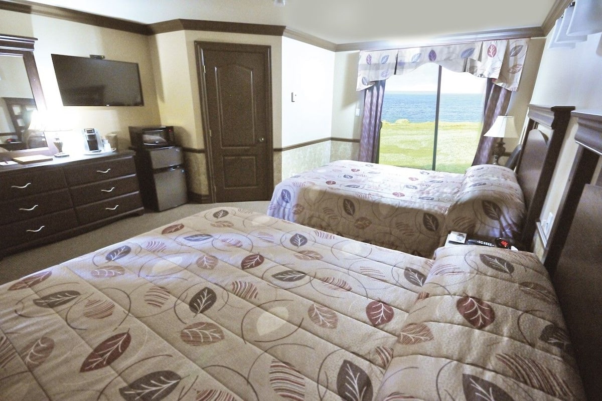 Quarto de hotel com duas camas, uma TV, uma escrivaninha com computador e vista para o mar.