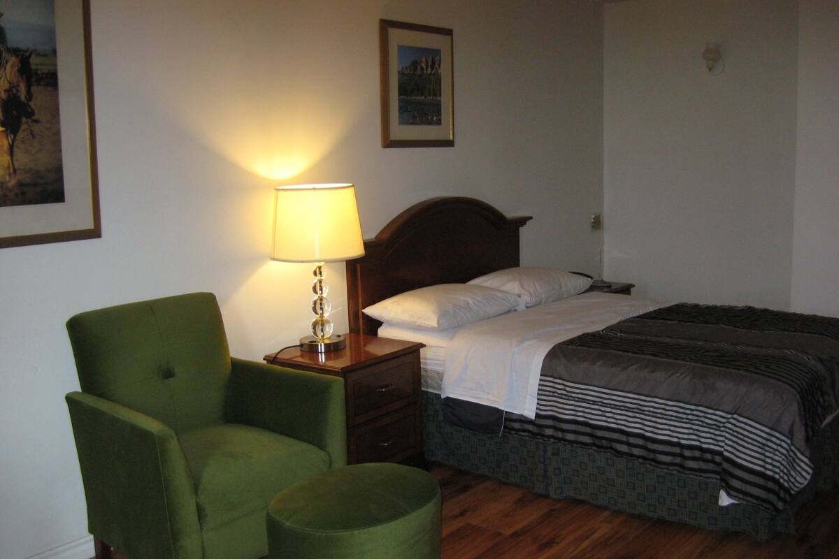 Quarto de hotel com uma cama, uma poltrona verde, uma mesa de cabeceira de madeira com abajur e um quadro na parede.