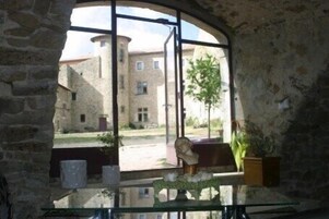 Quarto com parede de pedra, mesa de vidro com um vaso de flores e janela com vista para um edifício com uma árvore.