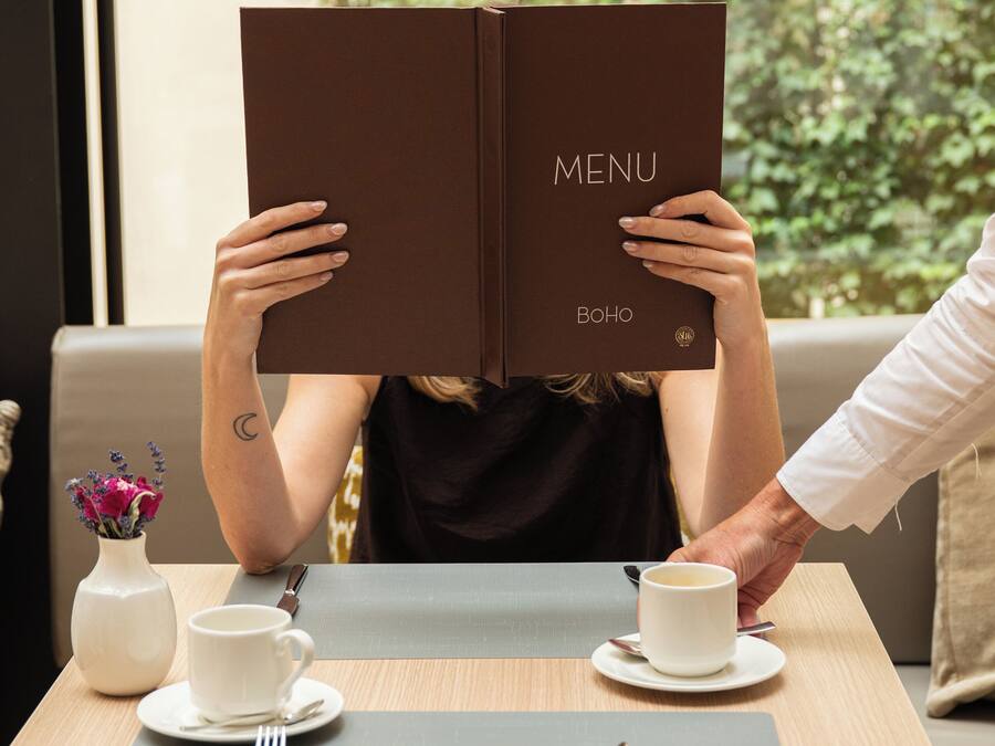 Uma pessoa segurando um cardápio à mesa, com uma xícara e pires, um vaso com flores e um garfo e colher sobre um guardanapo.