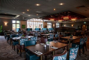Um restaurante espaçoso com várias mesas e cadeiras, janelas amplas e luminárias decorativas no teto.
