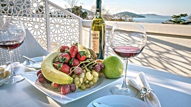 Mesa posta com uma garrafa de vinho, uma taça de vinho tinto, um prato de frutas e uma taça de vinho branco.