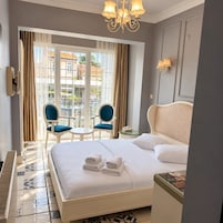 Quarto de hotel com uma cama grande, duas cadeiras, uma mesinha, um piano e vista para o exterior.