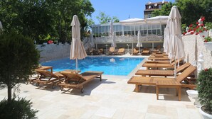 Uma área à beira da piscina com espreguiçadeiras e guarda-sóis, uma piscina azul cristalina e um edifício com grandes janelas ao fundo.