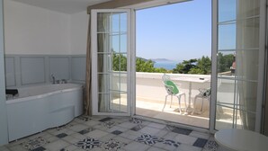 Um banheiro com banheira, vaso sanitário e chuveiro. Há uma grande porta de vidro deslizante que dá para uma varanda com vista para o mar e as montanhas.