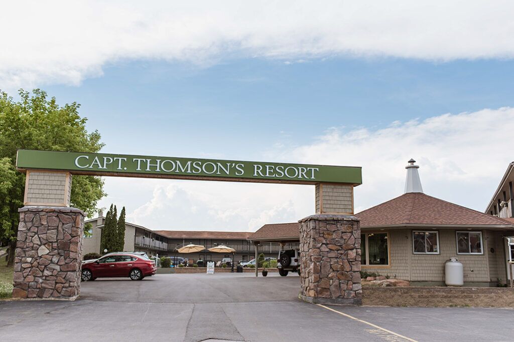 A entrada do Resort Capt. Thomson's, com um arco de pedra, um estacionamento e um edifício com telhado de azulejos.