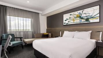 Quarto de hotel com uma cama grande, uma escrivaninha, uma cadeira, uma janela com cortinas e uma pintura abstrata pendurada na parede.