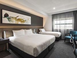 Quarto de hotel com uma cama grande, uma cama menor, uma escrivaninha, uma cadeira e uma janela com cortinas.