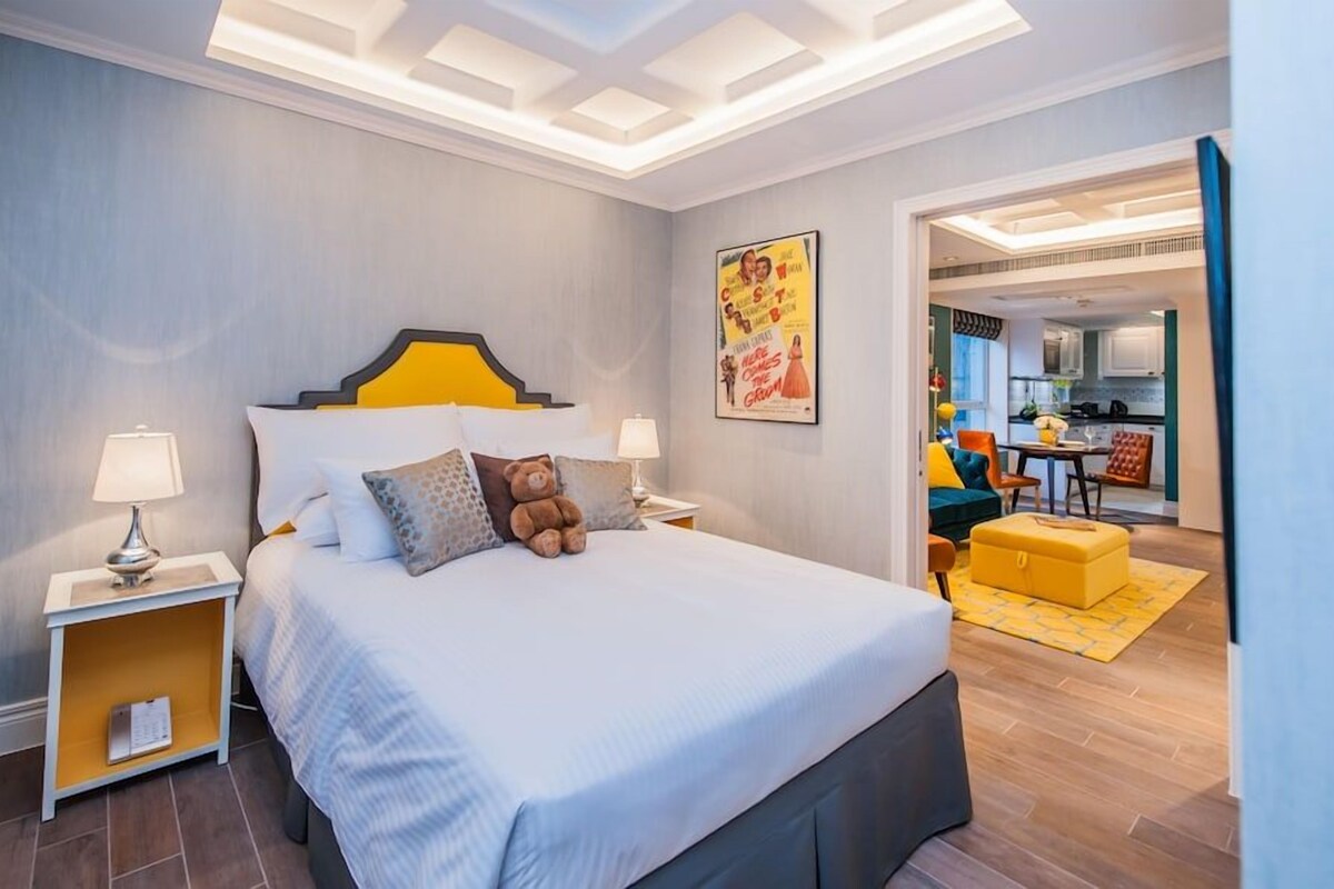 Quarto de hotel com cama, mesas de cabeceira, abajur, um urso de pelúcia, um cartaz e vista para a cozinha.