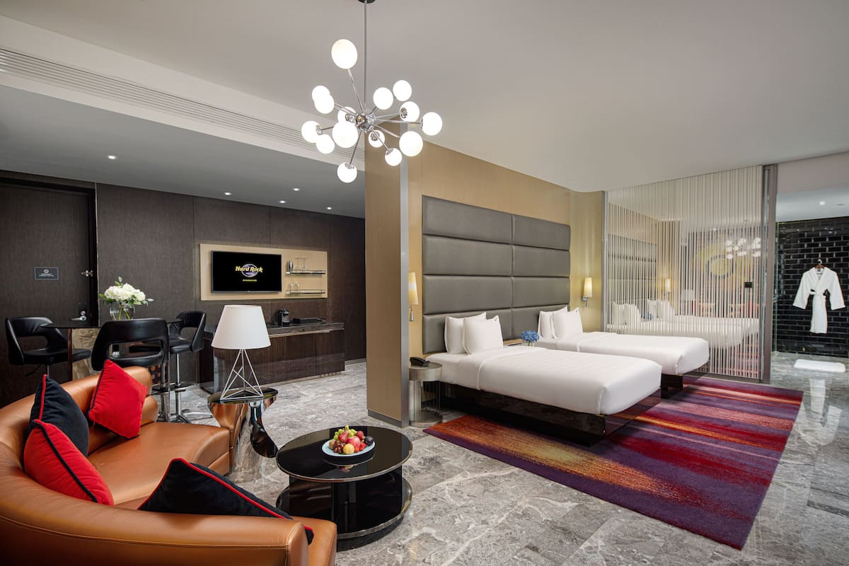 Quarto de hotel moderno com sofá, mesa de jantar, televisão, cama e banheiro visível por uma divisória de vidro.