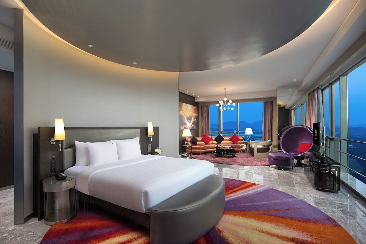 Quarto de hotel moderno com uma cama grande, área de estar com sofá e poltronas, televisão e vista para a cidade.