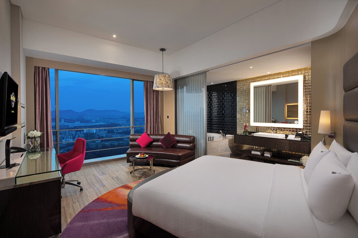Quarto de hotel com uma cama grande, uma escrivaninha, uma cadeira, um sofá, um espelho e vista para a paisagem urbana.