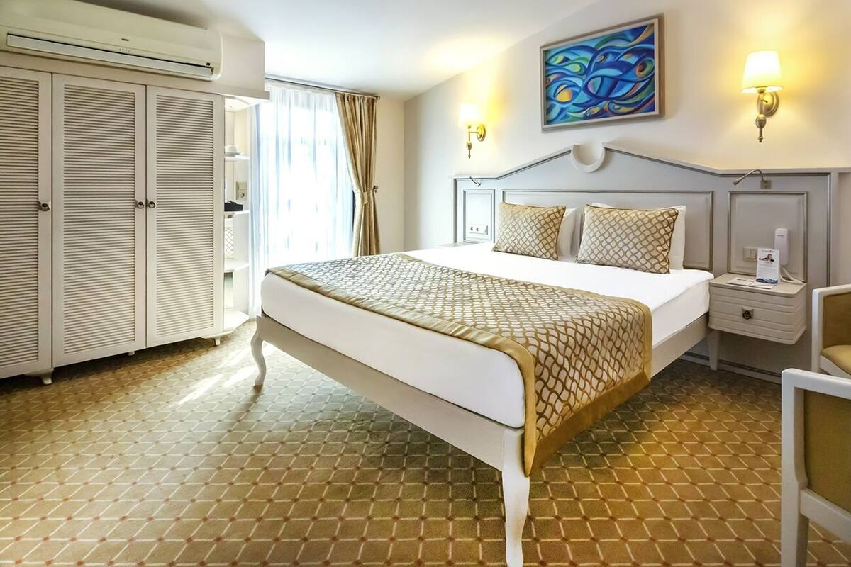 Quarto de hotel com cama, mesa de cabeceira, luminária, guarda-roupa e um quadro na parede.