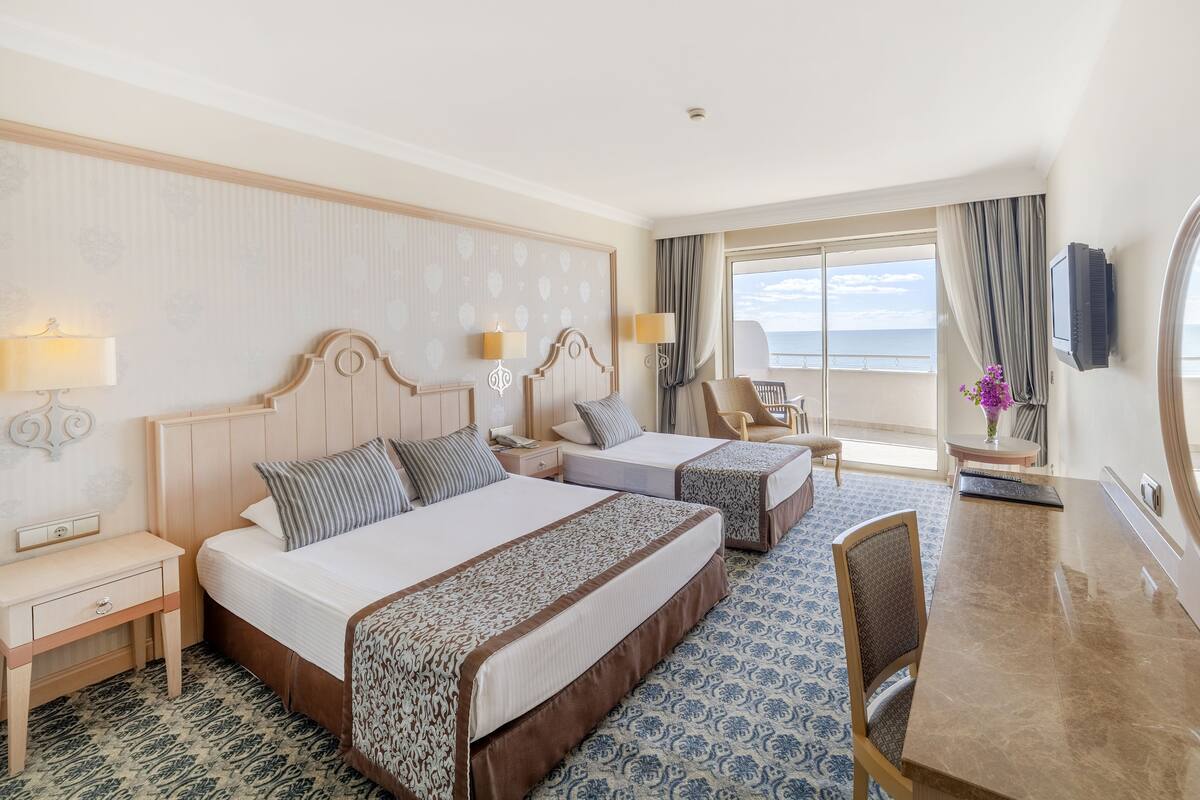 Quarto de hotel com duas camas, uma escrivaninha, uma cadeira, televisão e uma janela grande com vista para o mar.