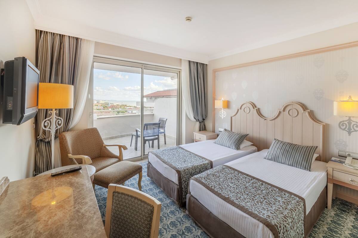 Quarto de hotel com duas camas, uma mesa, uma cadeira, uma TV e uma varanda com vista.