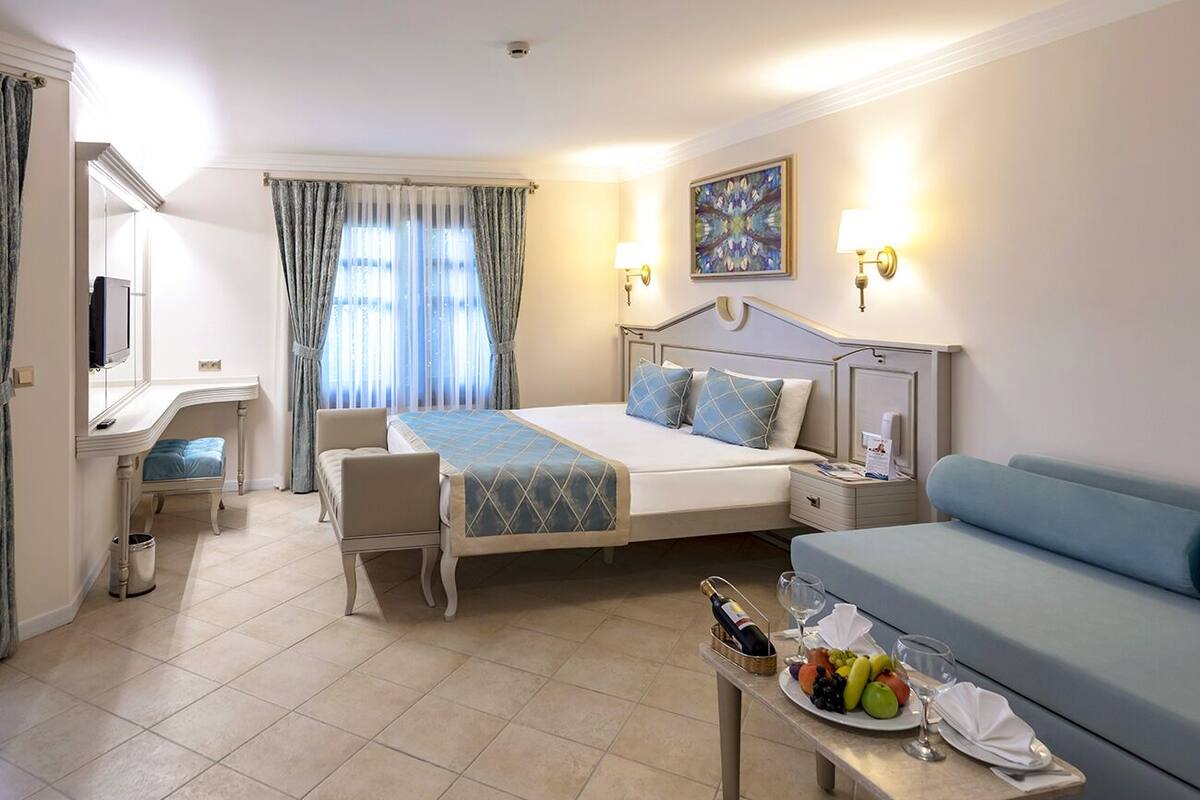Quarto de hotel com cama, sofá, escrivaninha e uma mesinha com garrafa e frutas.