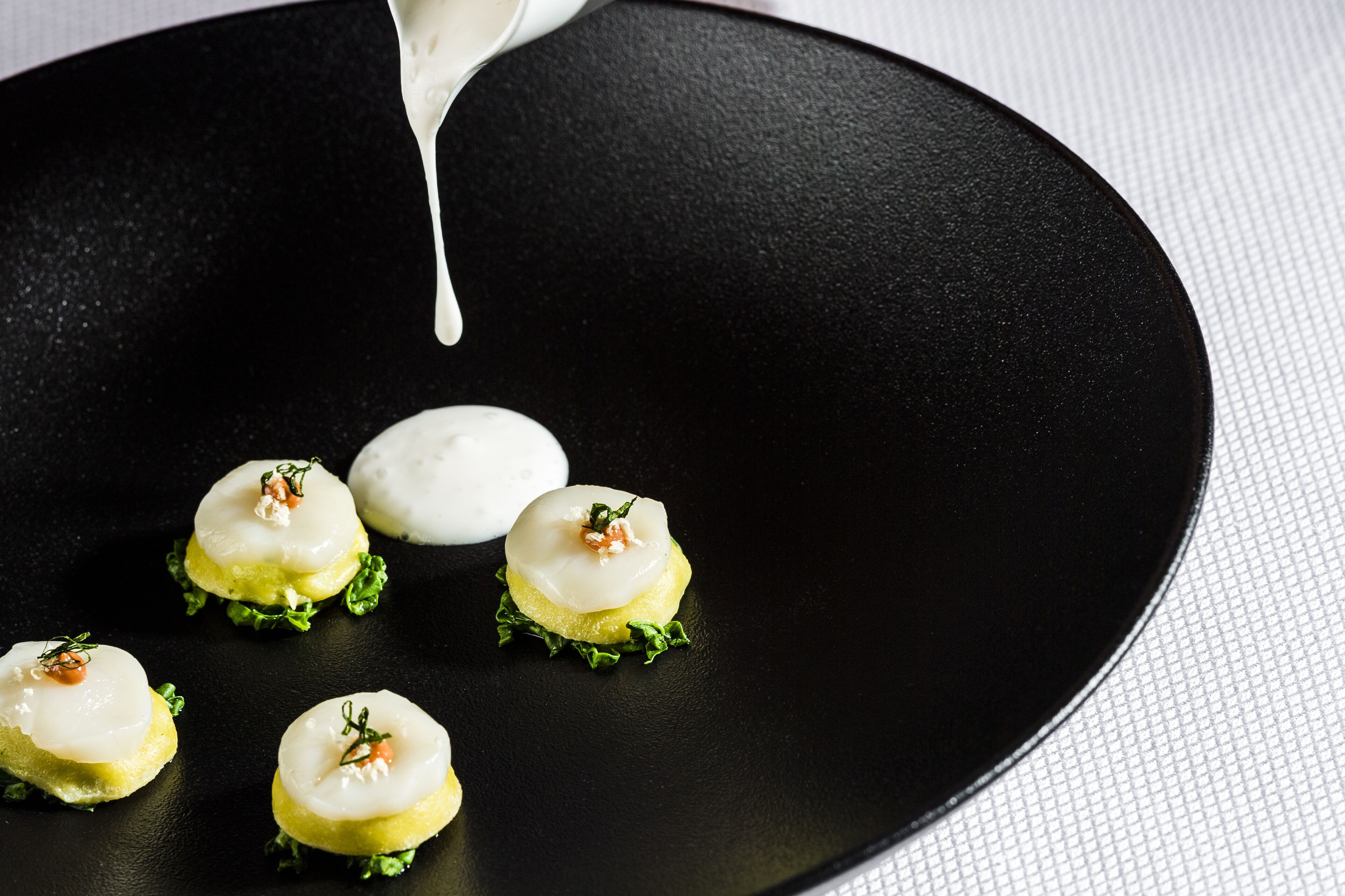 Um prato preto com quatro porções de comida, cada uma coberta com molho branco e decorada com uma pequena folha verde.
