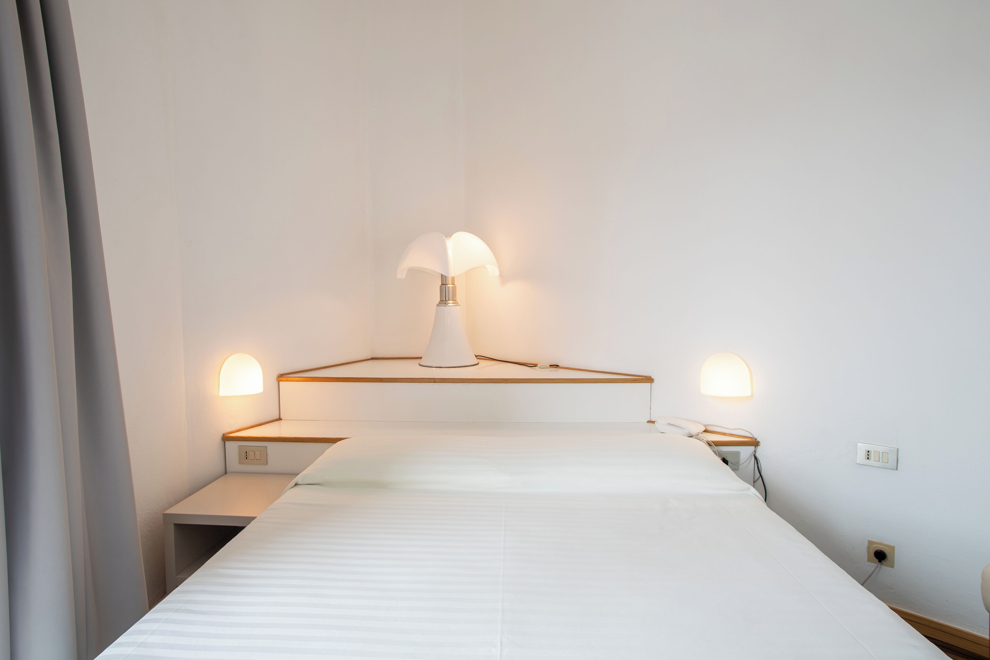 Quarto de hotel com uma cama grande, duas mesas de cabeceira, uma luminária fixada na parede e uma tomada.
