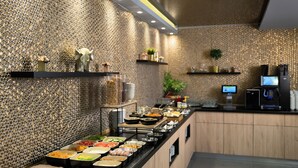 Uma área de buffet com diversas opções de comida, uma máquina de café e uma tela de televisão.