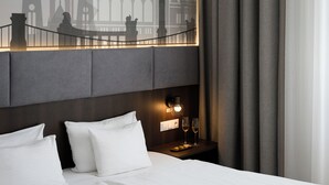 Quarto de hotel com uma cama grande, cabeceira com um mural da paisagem urbana, mesa de cabeceira com abajur e janela com cortinas.