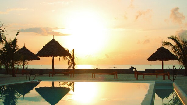 Piscina com vista para o pôr do sol, palmeiras e guarda-sóis de praia.