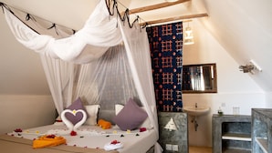 Um quarto com cama de dossel, mesa de cabeceira com toalha, criado-mudo, pequena mesa de cabeceira, prateleira na parede, espelho e um banheiro com pia e porta-toalhas.