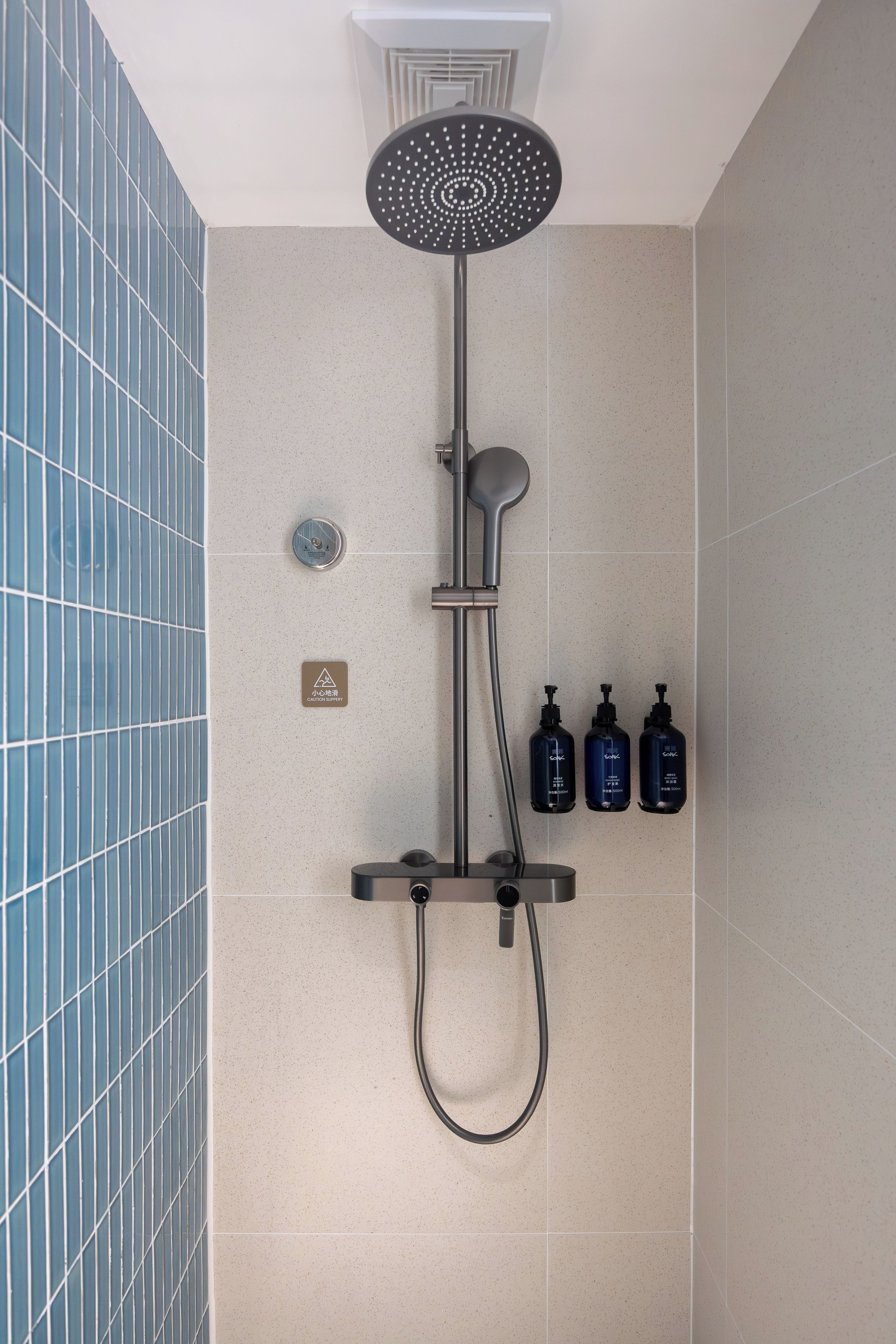 Chuveiro moderno com ducha de chuva, chuveiro manual e esfrega-espuma. Há três frascos de shampoo e condicionador na parede.