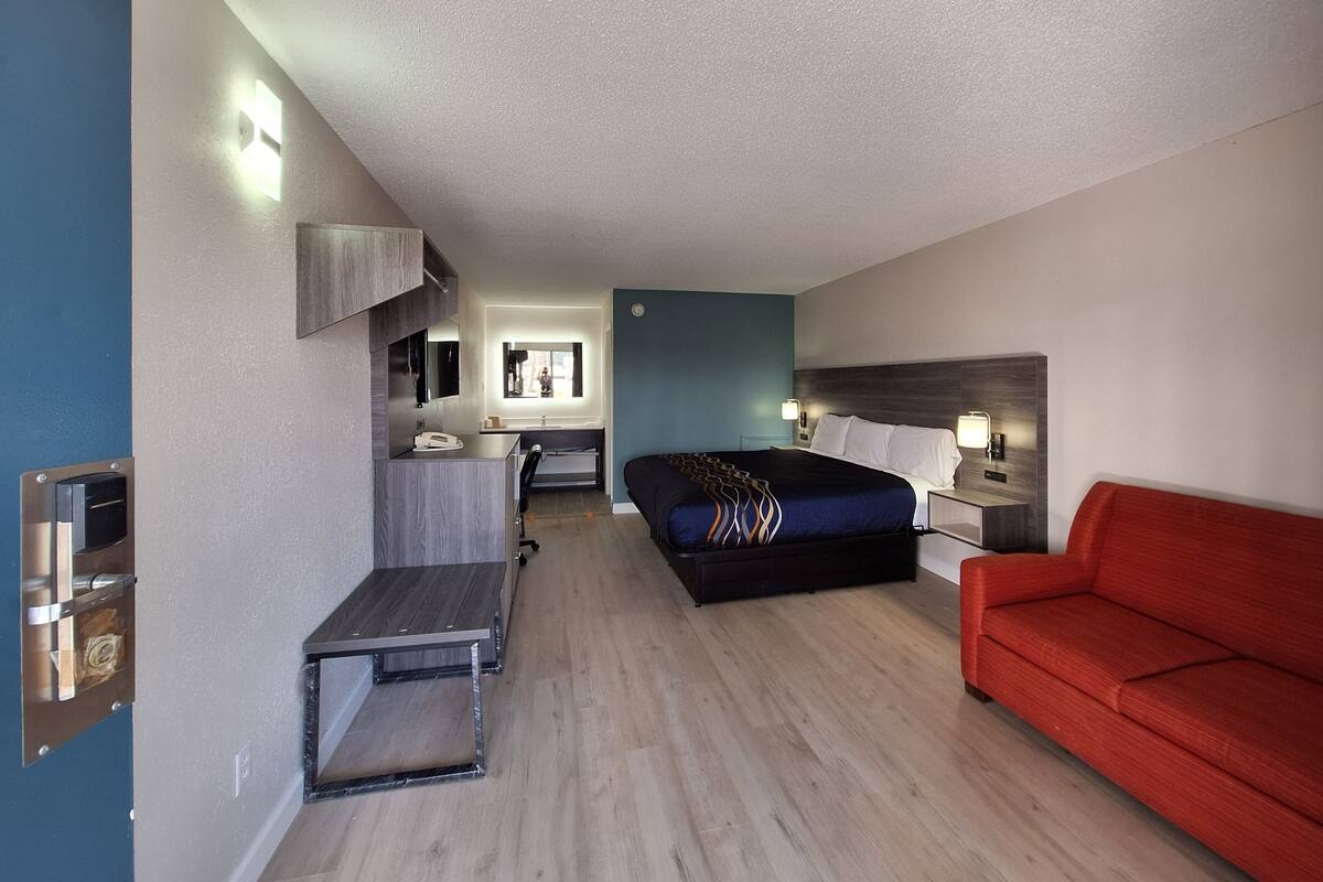 Quarto de hotel com cama, sofá vermelho, escrivaninha com telefone, espelho e janela.