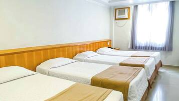 Quarto de hotel com três camas alinhadas, janela com cortinas e televisão presa na parede.