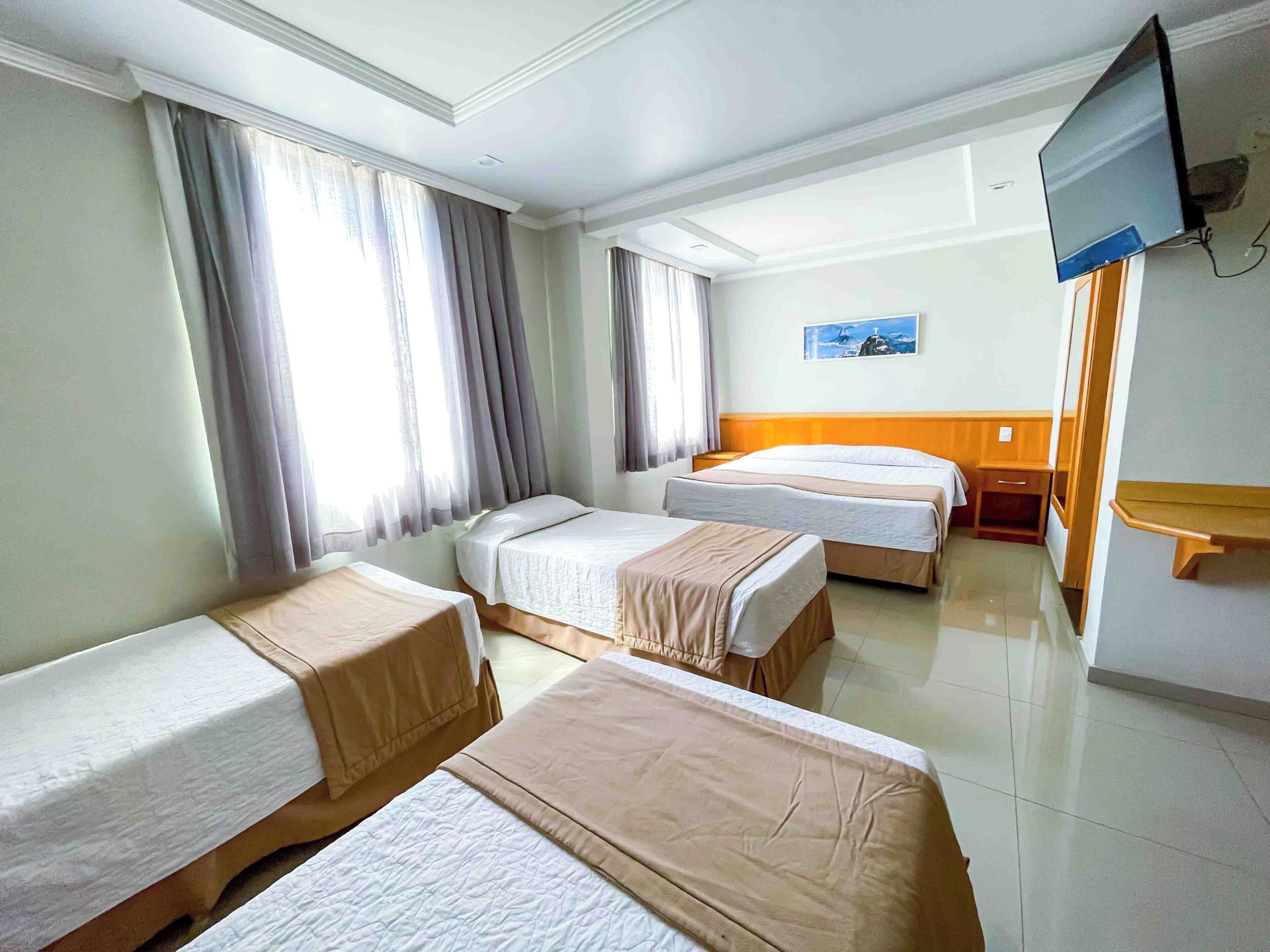 Quarto de hotel com três camas de solteiro, uma televisão presa à parede e uma janela com cortinas.