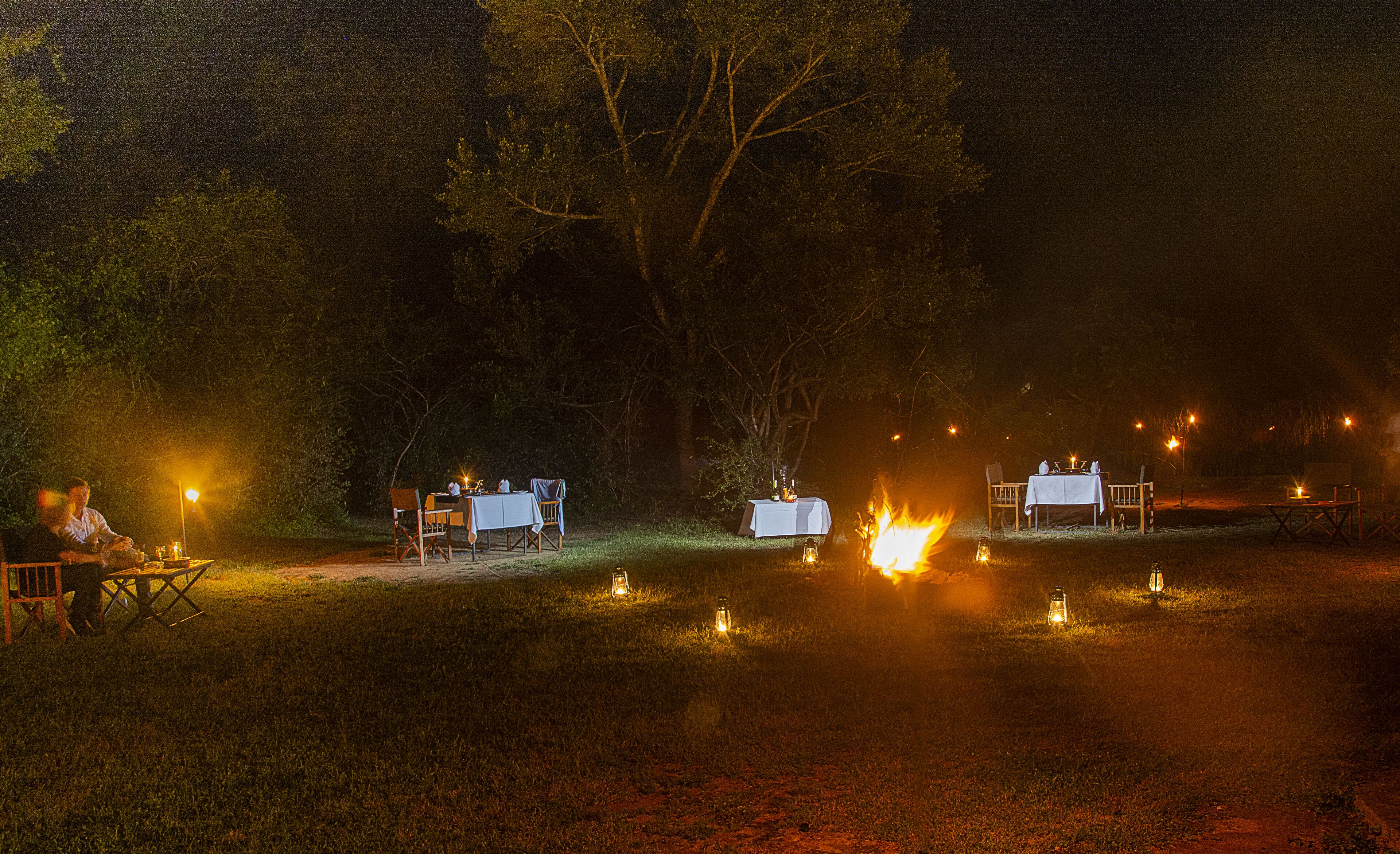 Uma área de refeições ao ar livre com mesas e cadeiras, cercada por árvores e iluminada por lanternas.