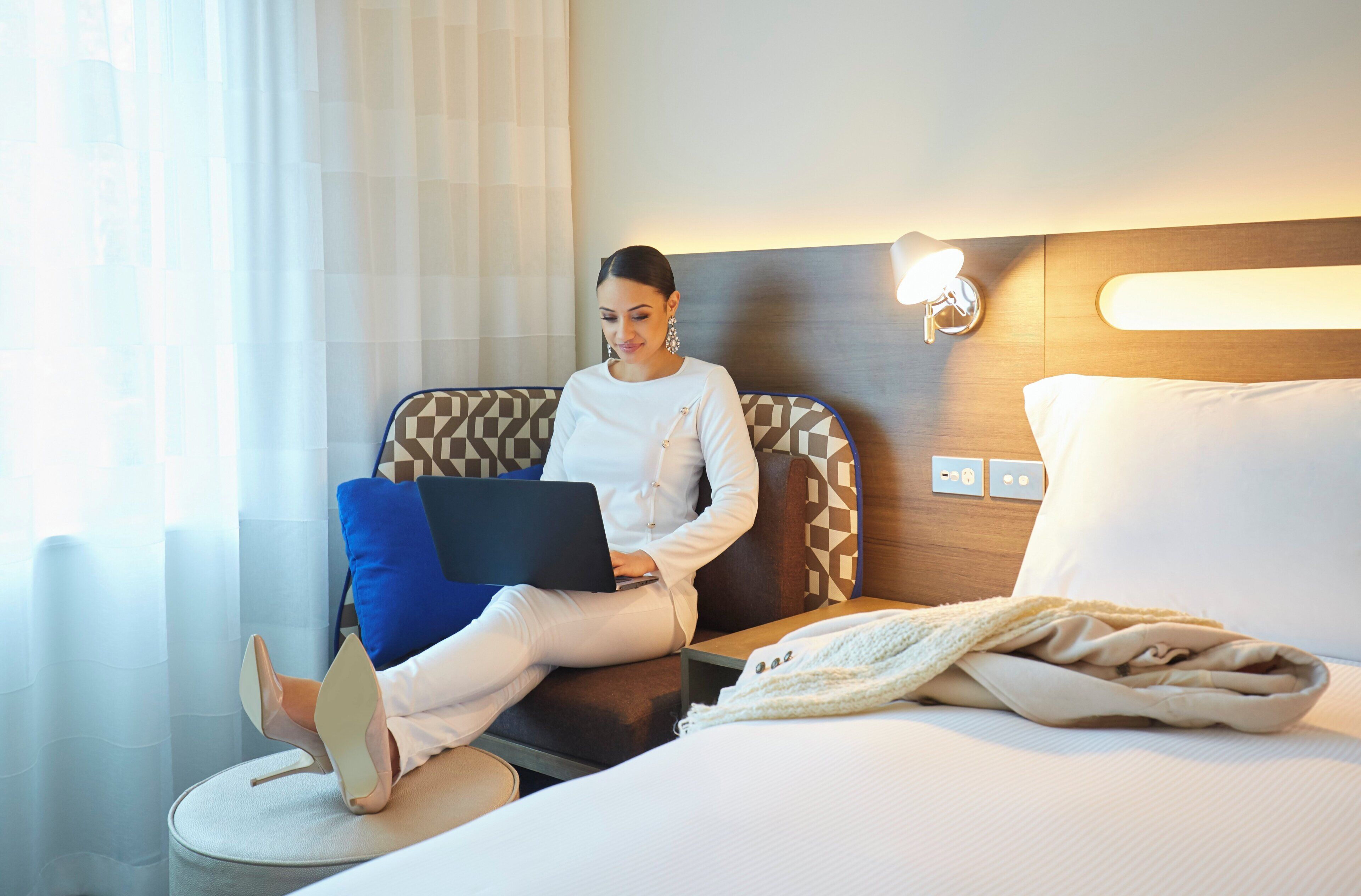 Uma pessoa sentada na cama com um laptop, um travesseiro e um cobertor.