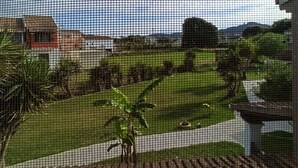 Vista pela janela, com chão de madeira, área gramada com um caminho e edifícios ao fundo.