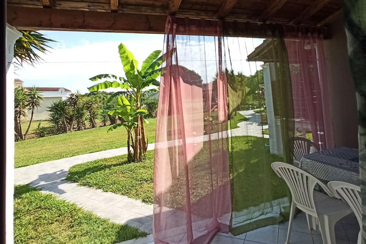 Um alpendre coberto com teto de madeira, uma mesa e cadeiras, e vista para um jardim com palmeiras.