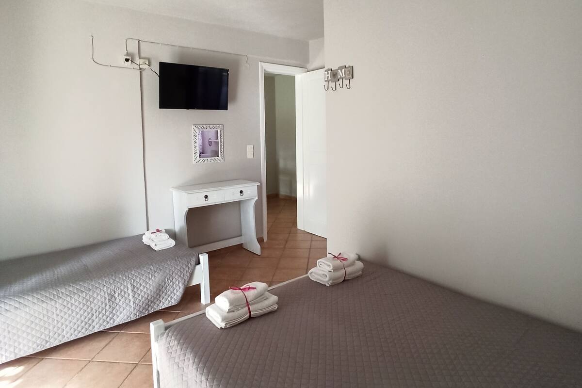Quarto de hotel com duas camas de solteiro, uma pequena escrivaninha, televisão de tela plana na parede e corredor que dá para outro quarto.